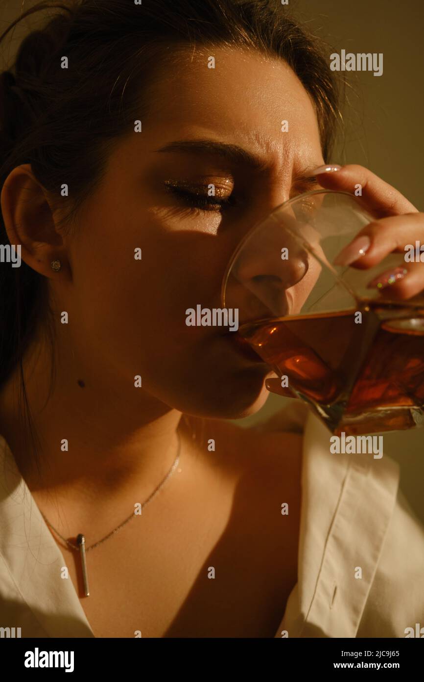 Young woman drinking alcohol from glass against sunny wall background ...