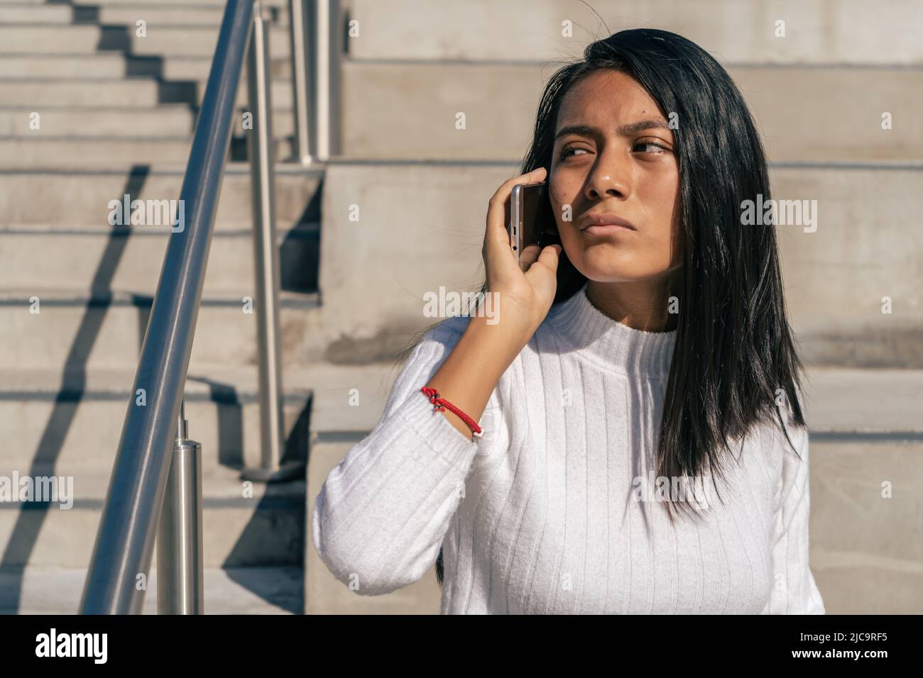 Young Latin woman talking on the phone sitting on a concrete ladder and ...