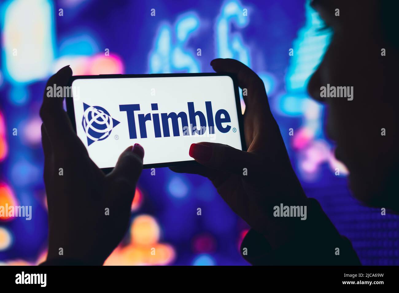 In this photo illustration, a silhouetted woman holds a smartphone with the Trimble Inc. logo ...