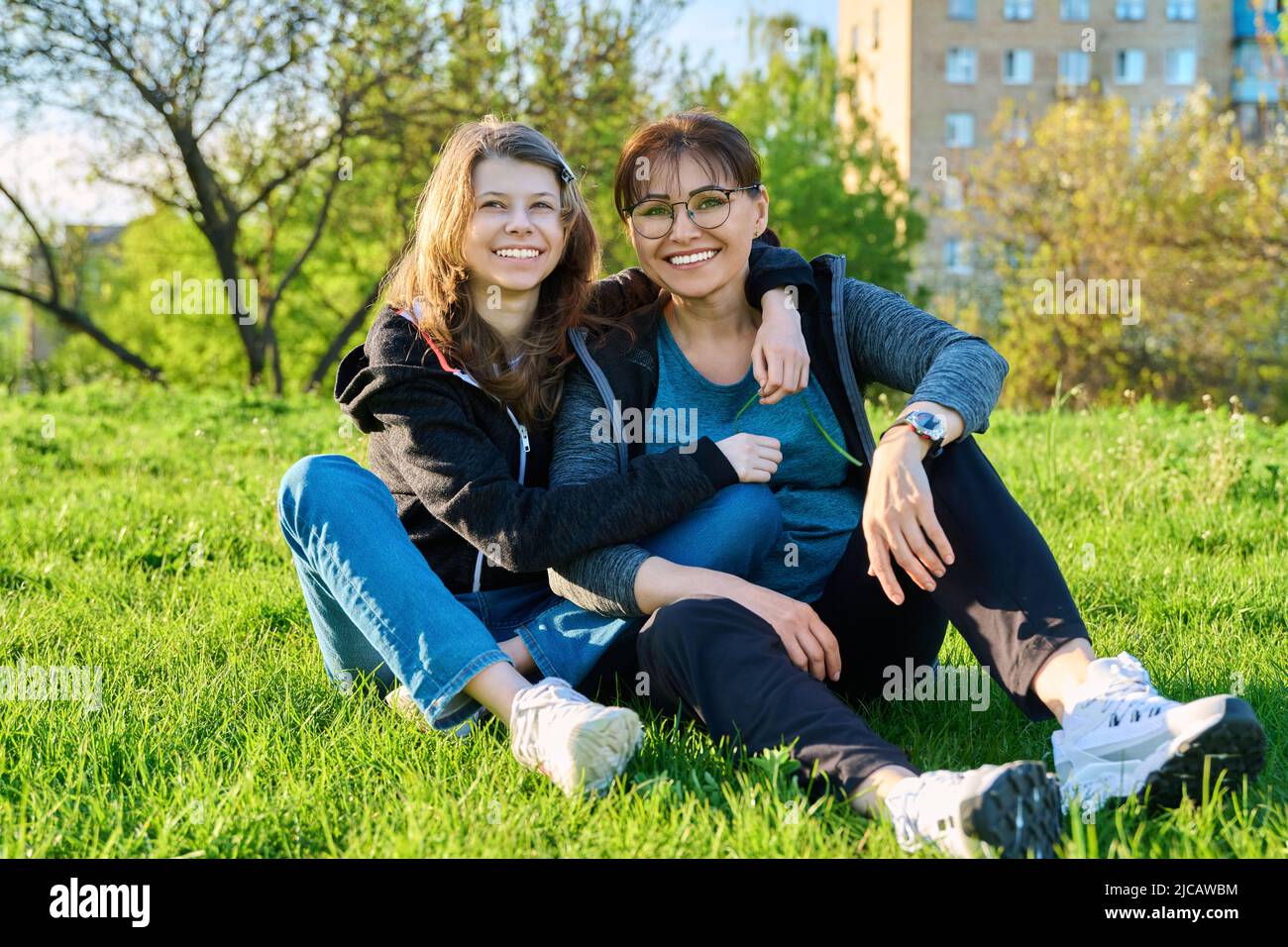 Happy mother and daughter hugging sitting on grass, looking at camera ...