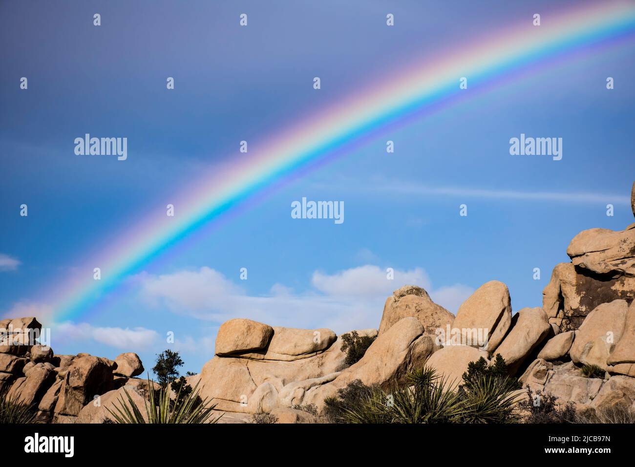 Rainbow arches across blue sky crossing desert landscape. Stacked rock ...