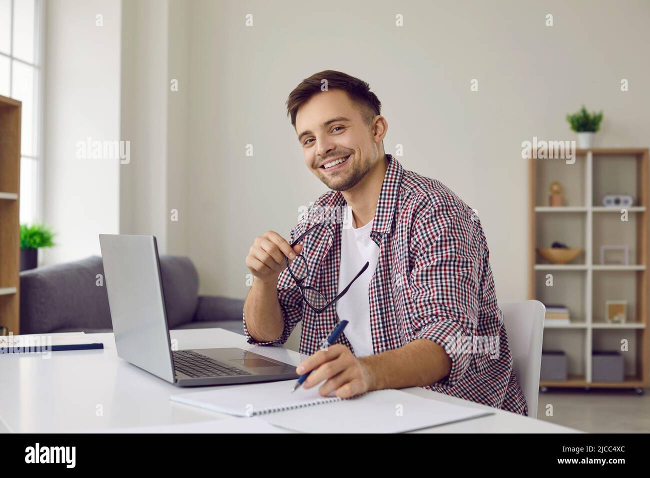 Happy student sitting at desk with laptop and notebook, holding glasses ...