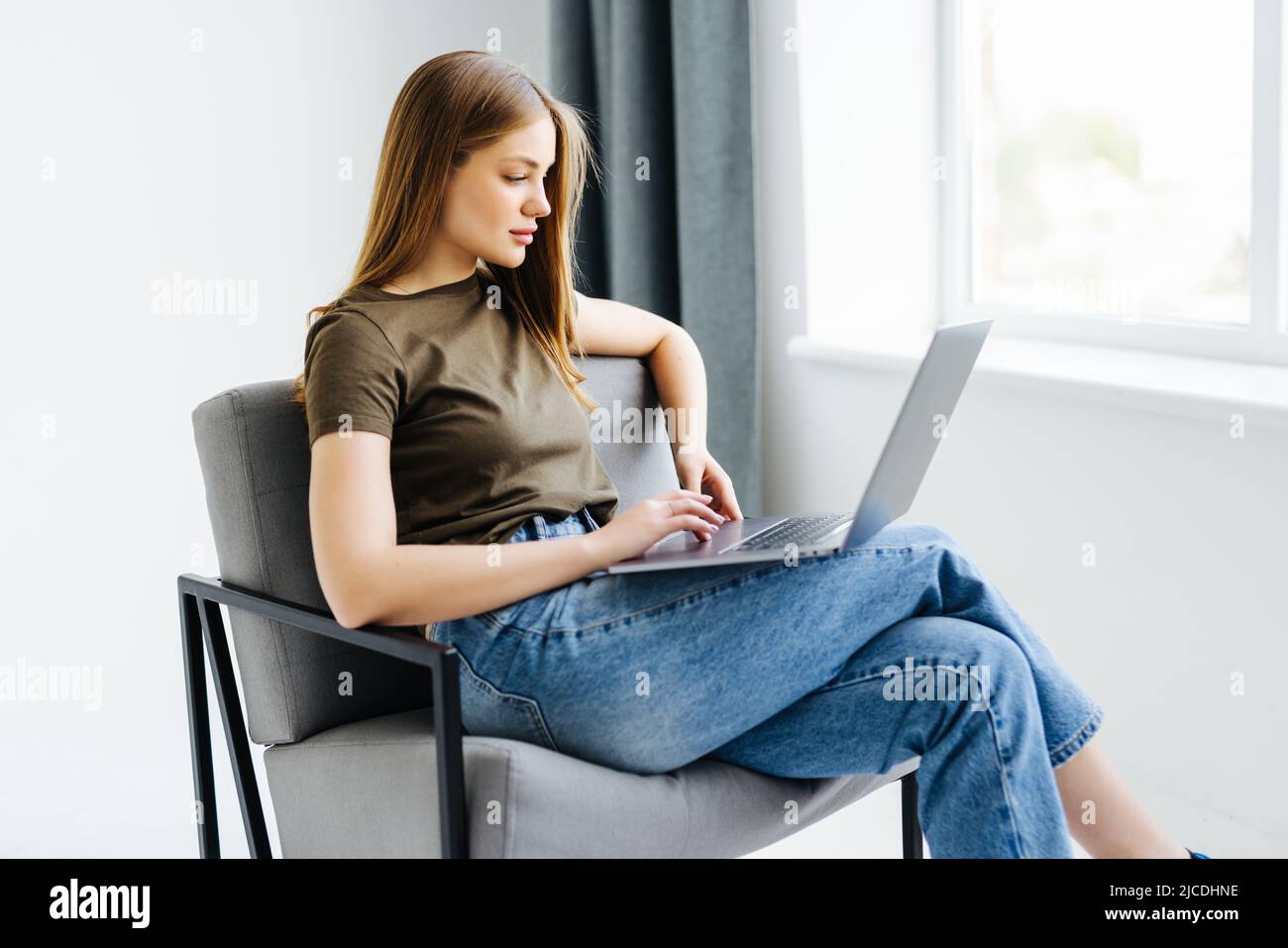 Young smiling brunette girl is sitting on modern chair near the window ...