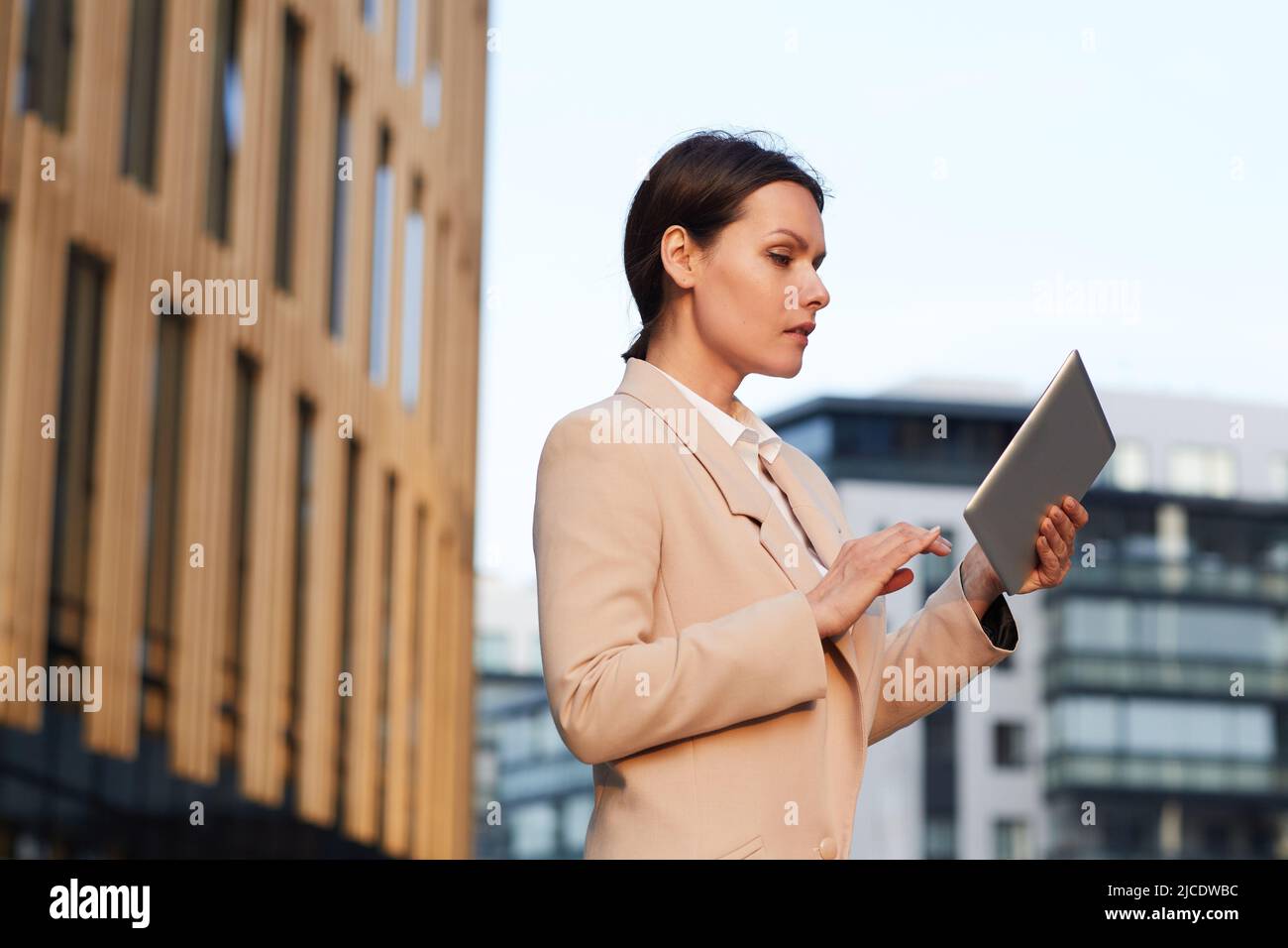 Thoughtful busy lady in light jacket standing outdoors and working with ...