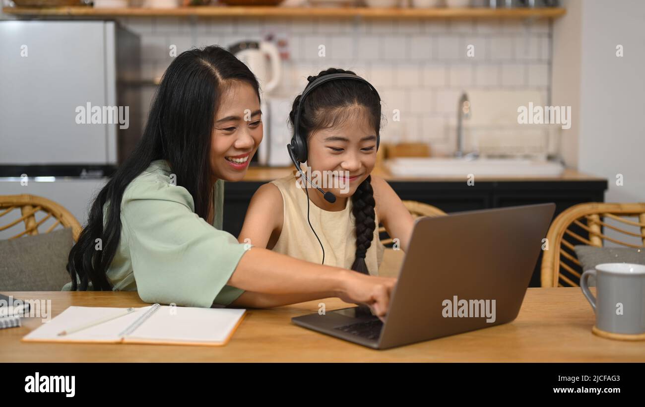 Smiling Asian mother helping her daughter doing homework during studying through online learning ...