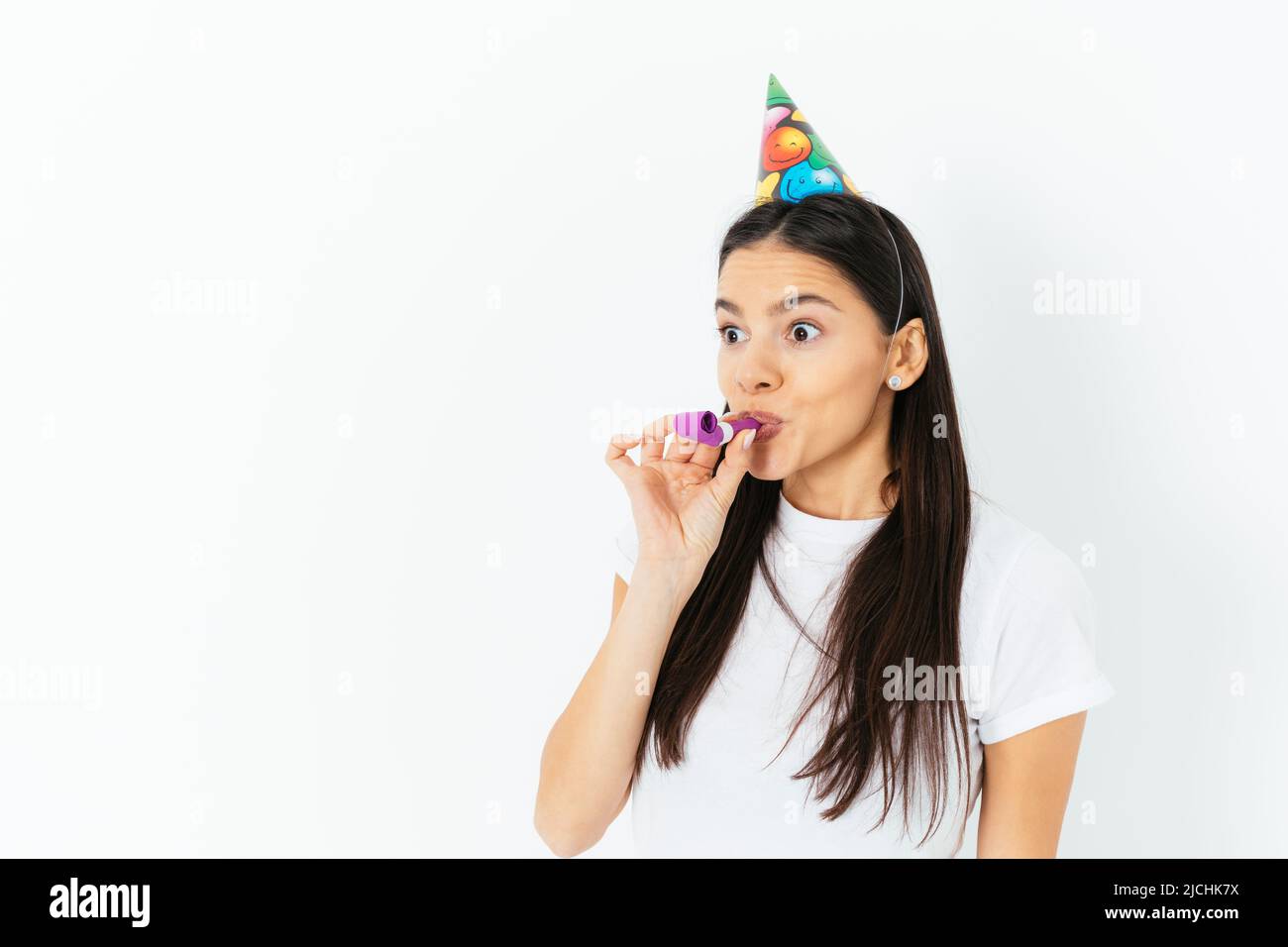 Portrait of funny young woman wearing birthday cap blowing kazoo pipe ...