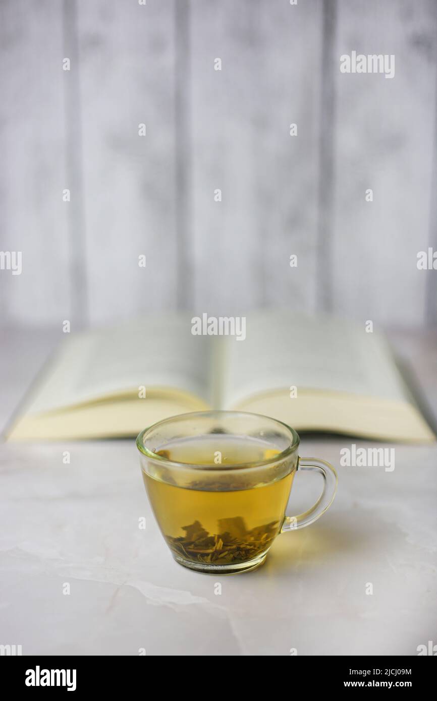 Brewed herbal green tea in a transparent cup in front of an open book ...