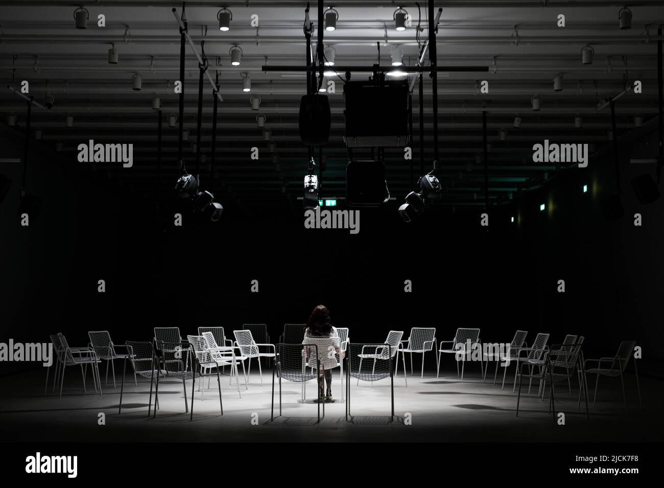 A girl sits in a circle of chairs under a beam of light in a dark room ...