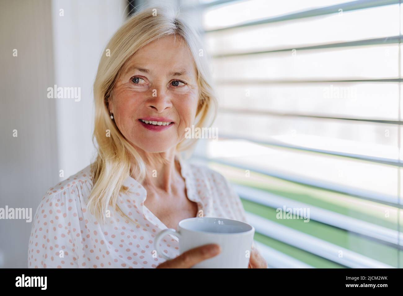 Attractive senior woman drinking tea and standing by window at home ...