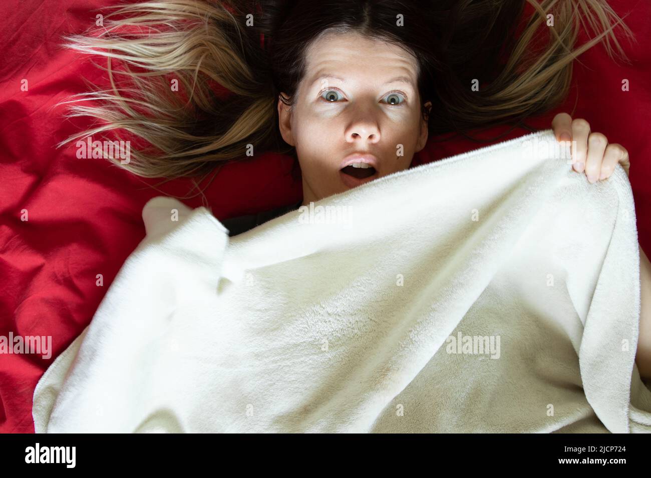 A young girl looks with surprise in bed on a red sheet and is covered ...