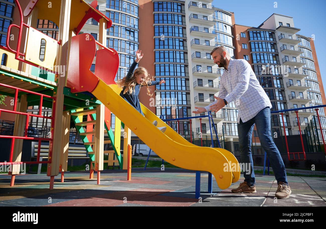 Handsome dad catching cheerful daughter, riding on slide at modern ...