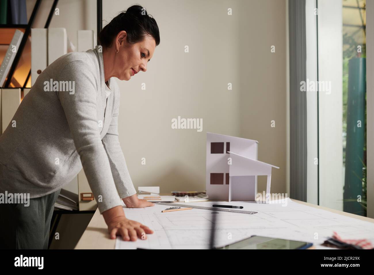 Pensive mature female architect leaning on table with building ...