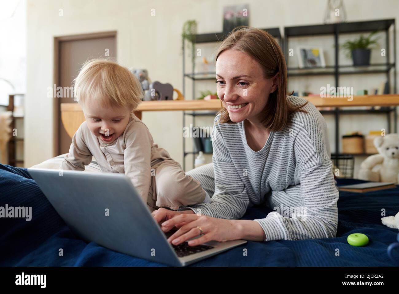 Smiling young mother typing on laptop and doing her online work while ...