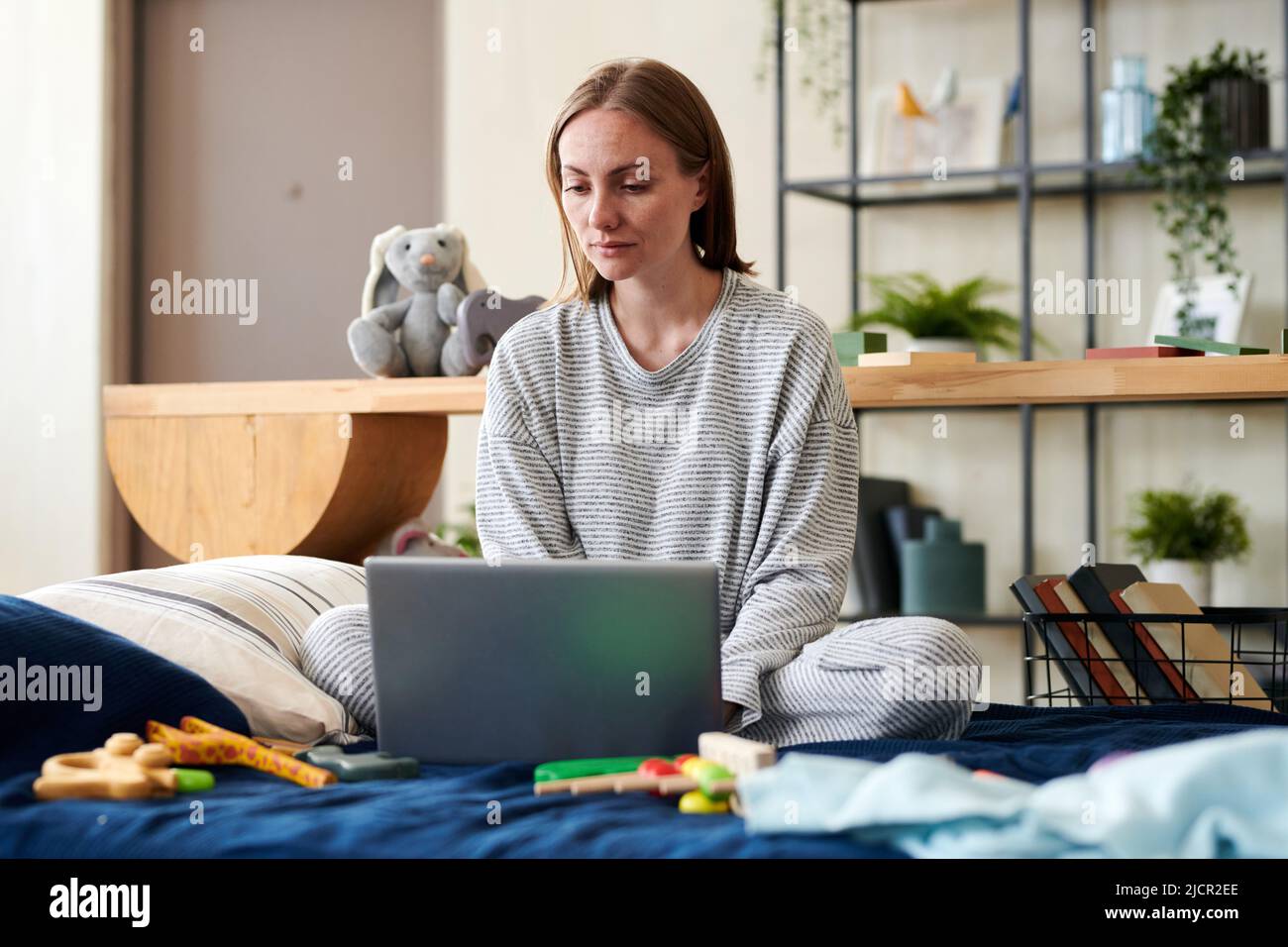 Young mother using laptop while sitting on bed among toys during sleep ...