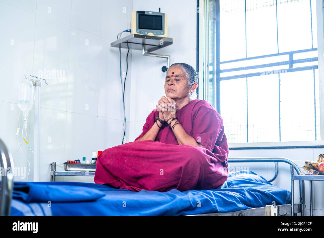 Senior woman in operation gown at hospital praying god by joining hands ...
