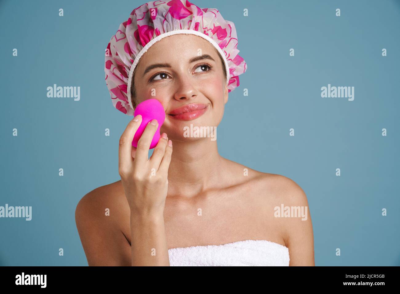 Young woman wearing shower cap smiling while using cosmetic sponge ...