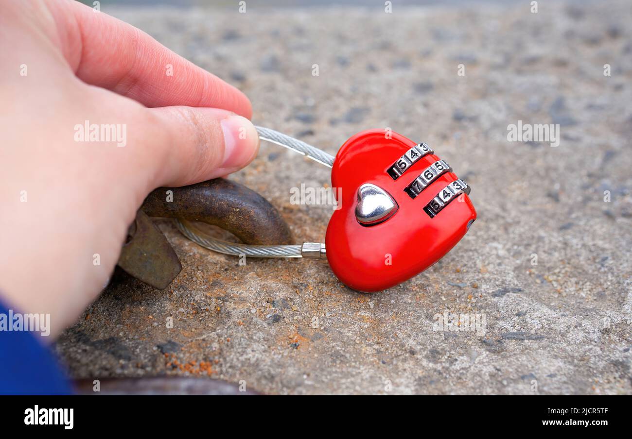 Fingers holding a red heart shaped combination lock against a concrete ...