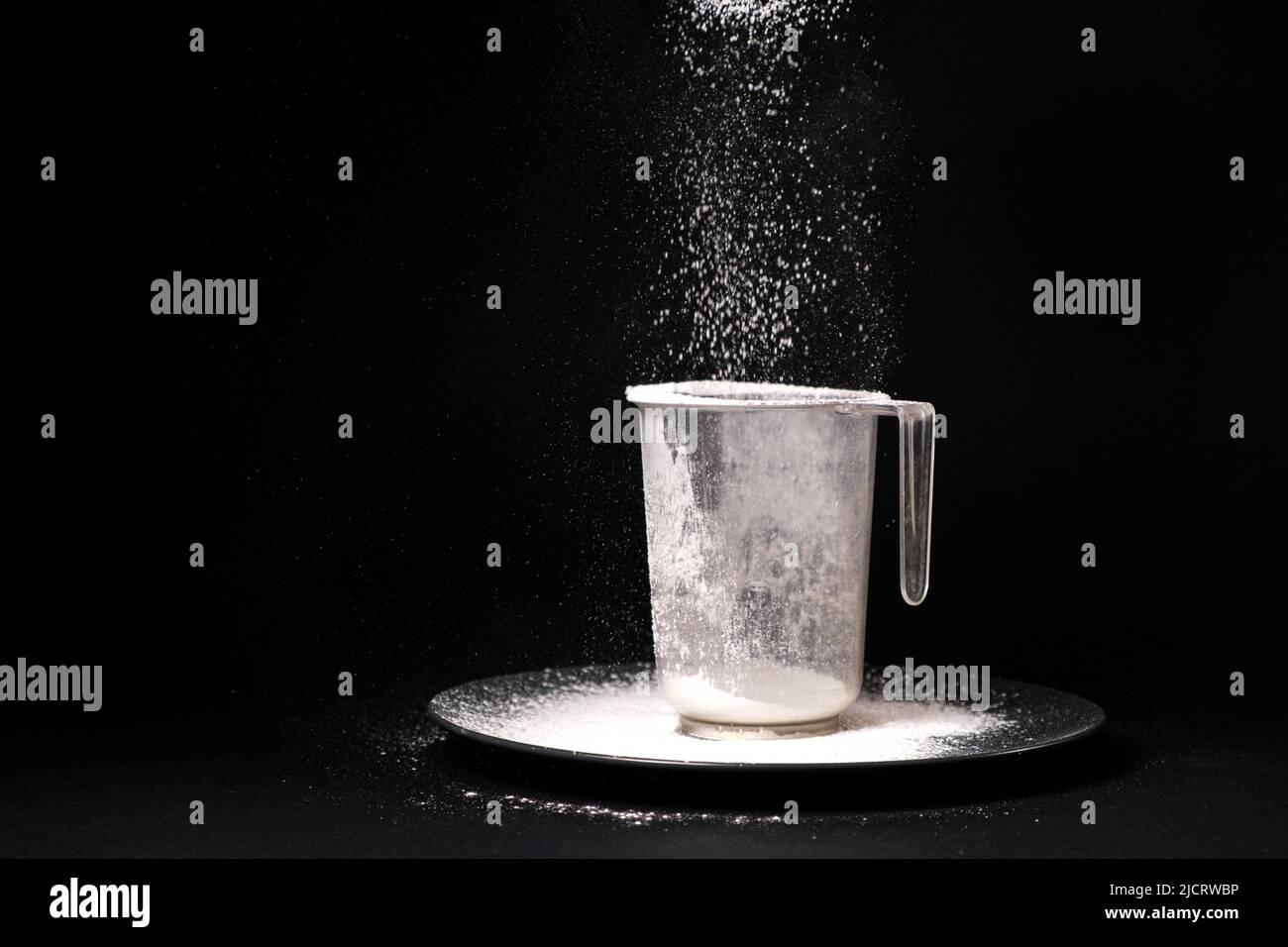 White wheat flour is poured into a measuring cup on a black background ...