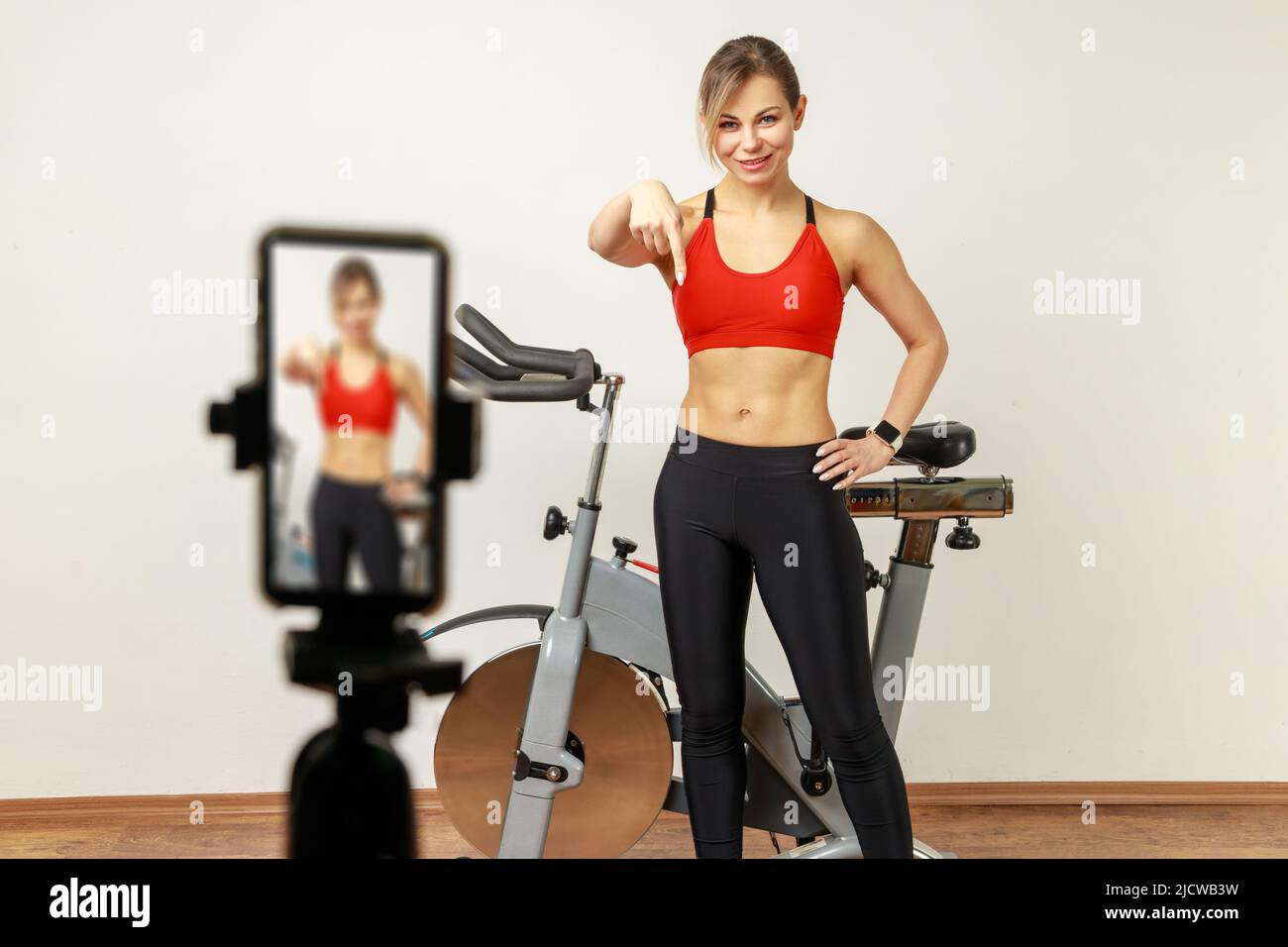 Smiling sporty woman blogger in top standing near exercise bike ...