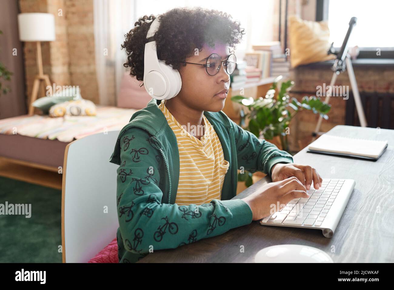 African little boy in wireless headphones typing on keyboard at table ...