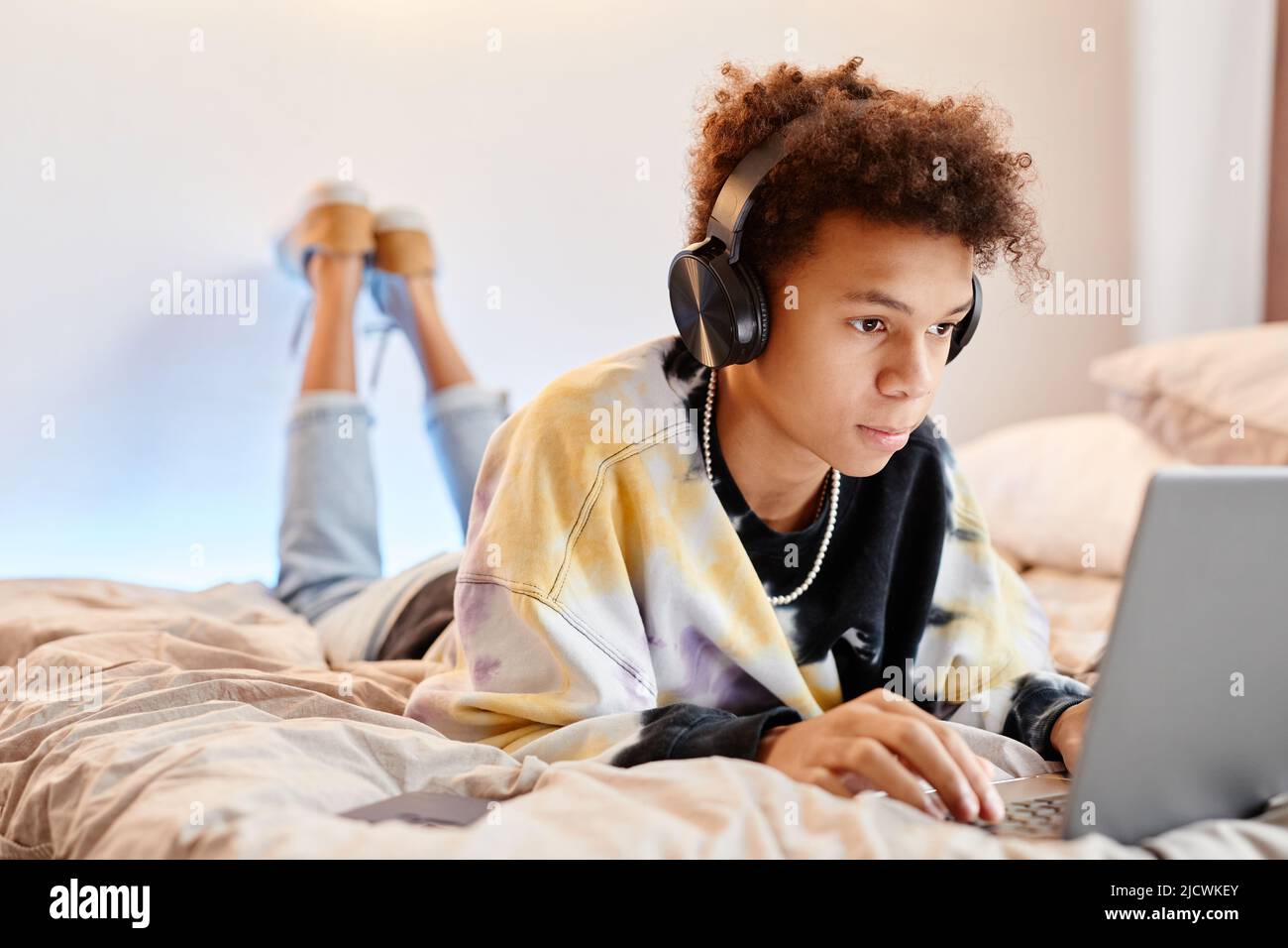 Portrait of teen black boy using computer while lying on bed in minimal ...