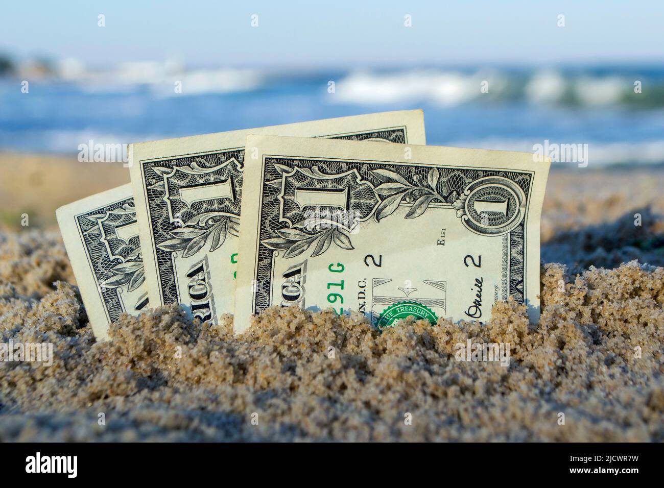 Three one dollar banknotes dug halfway into sand on sea beach on sunny ...