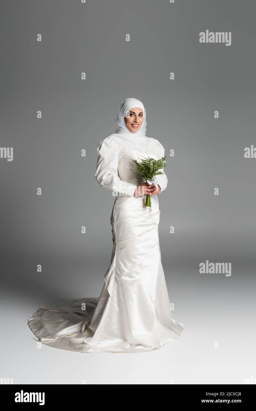 full length of pleased muslim bride in white dress and hijab holding ...