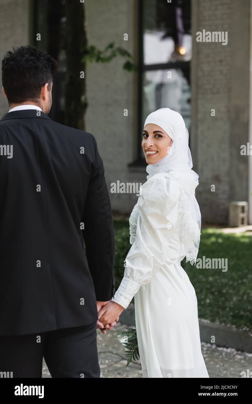 groom holding hands with cheerful muslim bride in wedding dress and ...