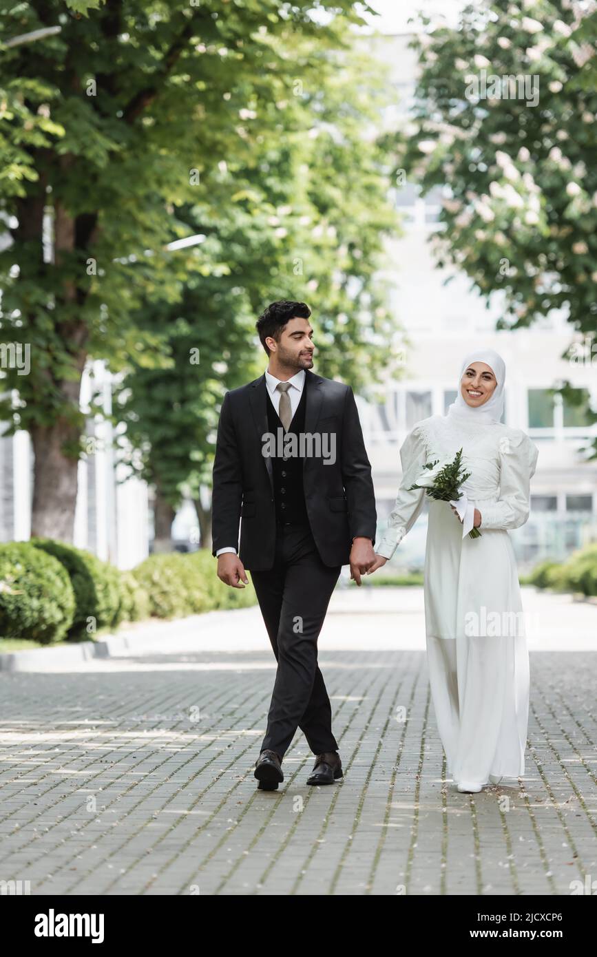 happy groom holding hands with smiling muslim bride in hijab with ...