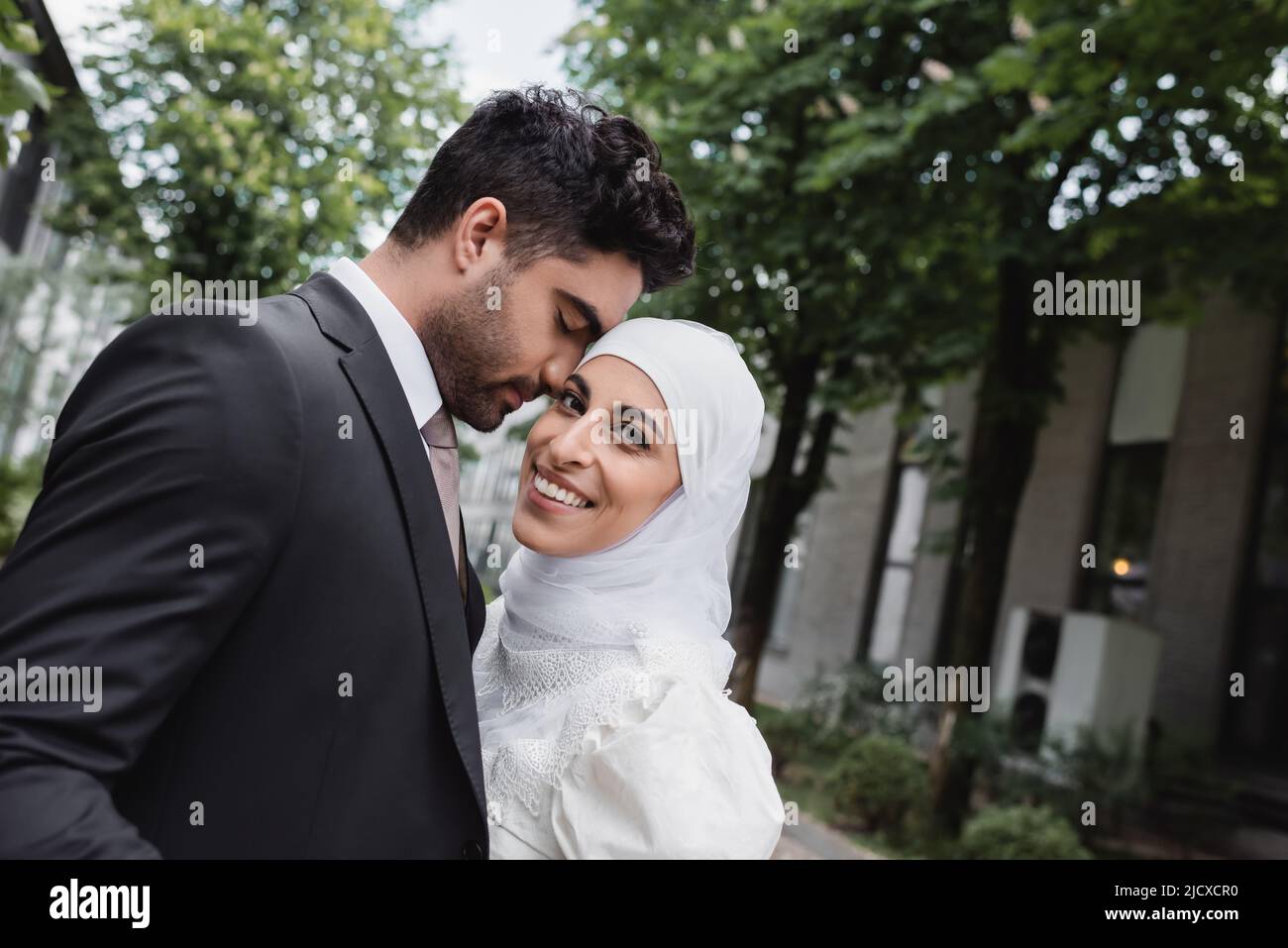 groom in suit hugging cheerful muslim bride in hijab and white dress ...