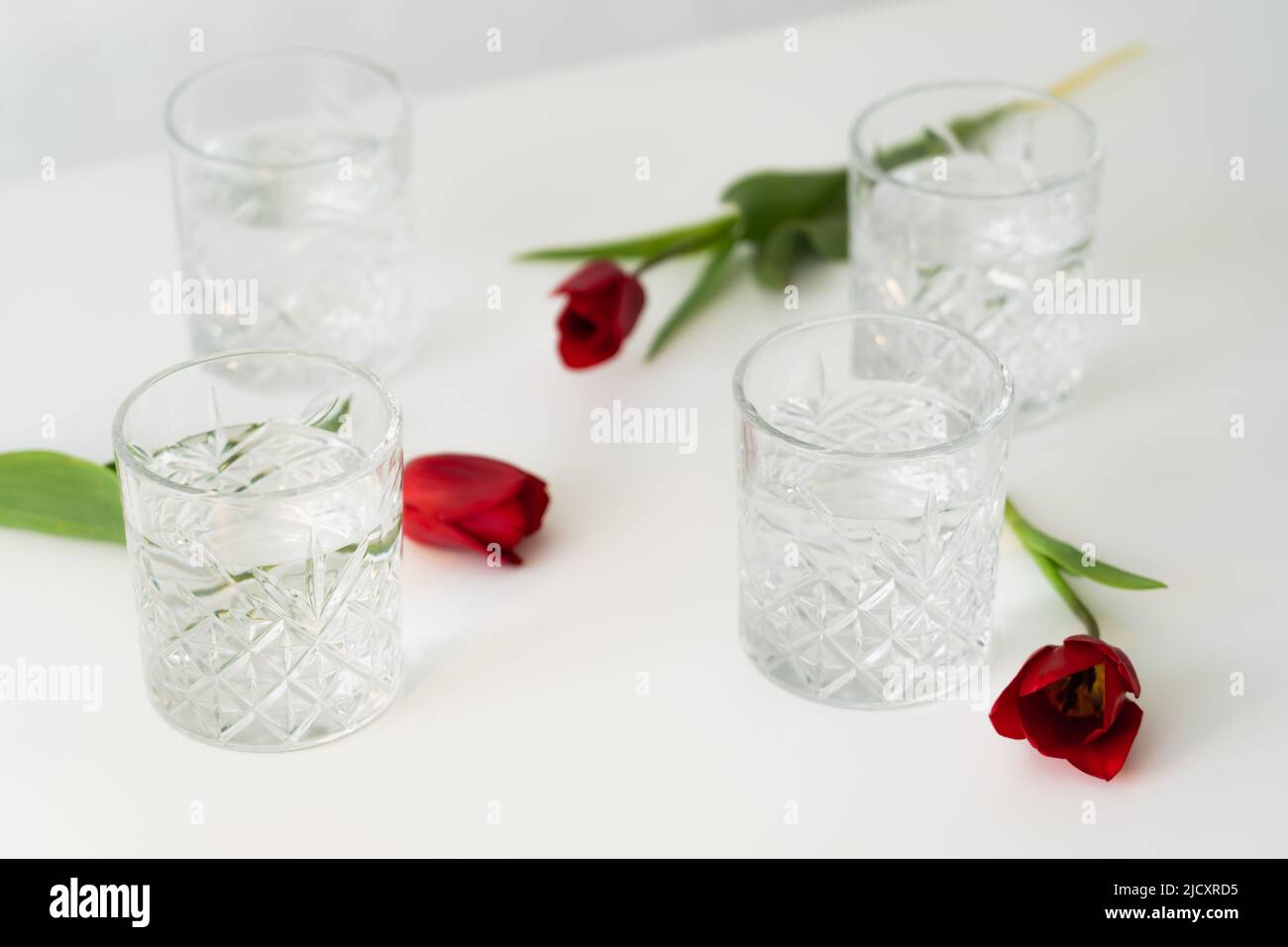 faceted glasses with water near red tulips on white blurred background ...