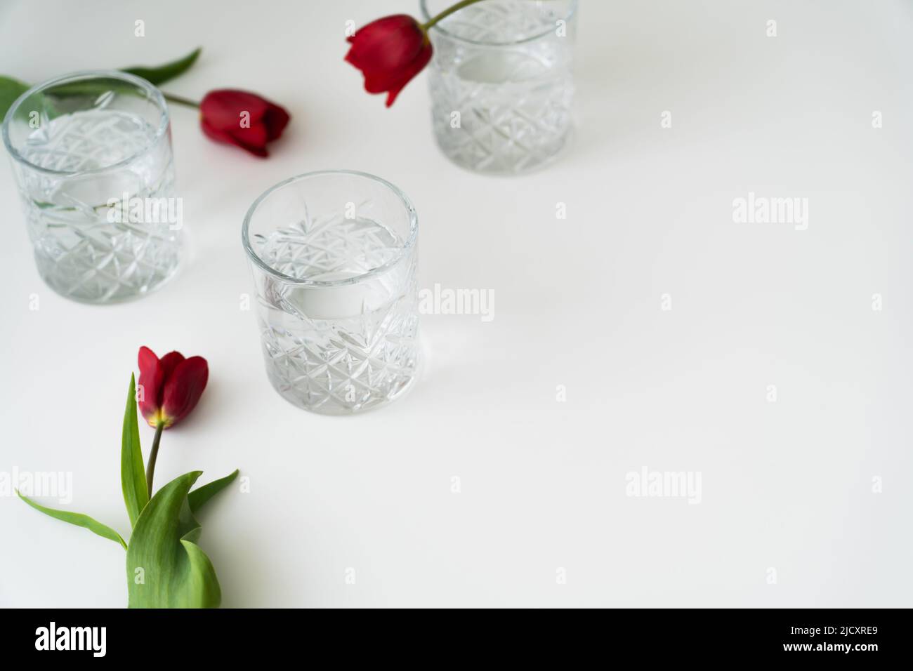 glasses with faceted pattern and clean water near red tulips on white ...
