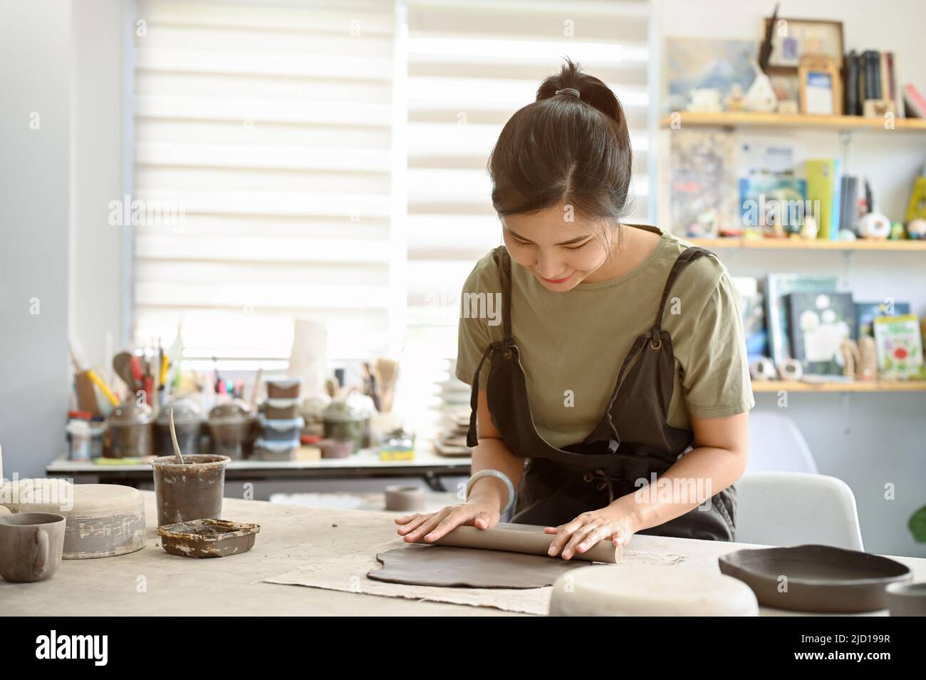 Relaxed and calm millennial asian woman rolling clay on worktable with ...
