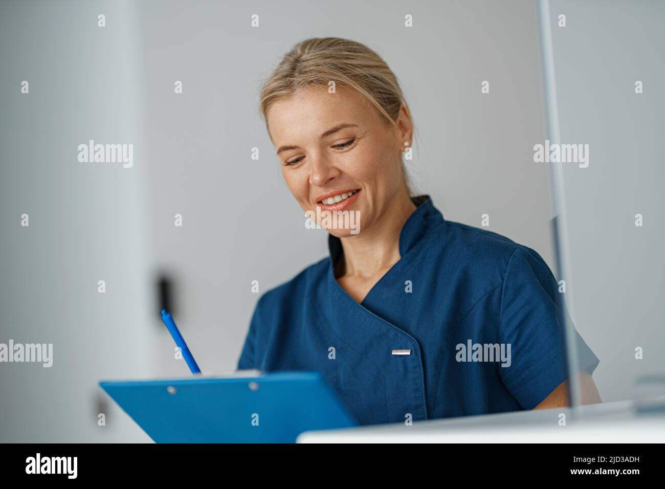 Smiling doctor making notes in clipboard standing near reception in ...