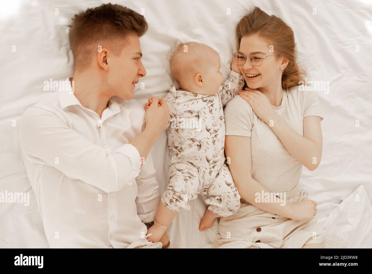 Young mother and father lay with little baby on bed, have fun and enjoy ...
