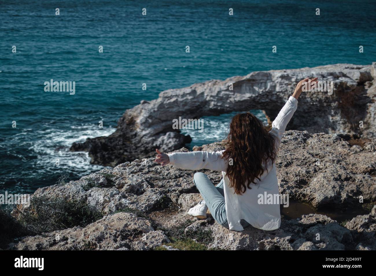 Carefree woman from behind sit at the edge and looking at sea cave arch ...