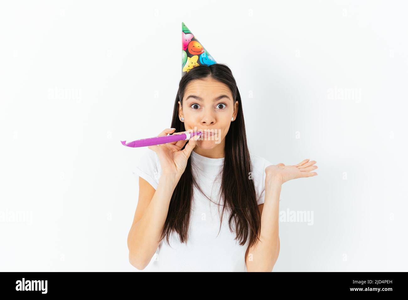 Cheerfully surprised young woman in festive birthday cap, blowing kazoo ...