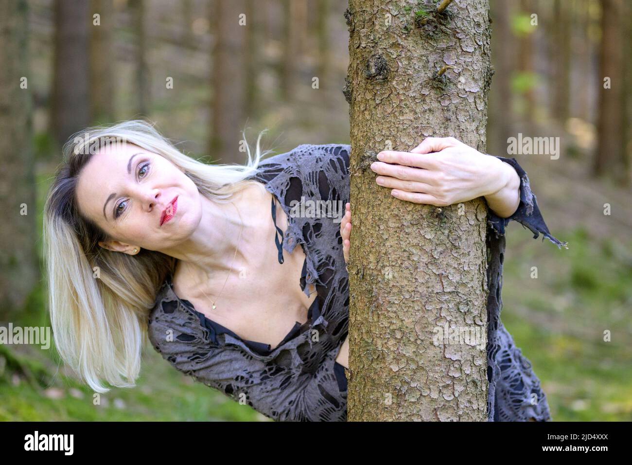 Playful blond woman peering around a tree trunk with a cheeky grin ...
