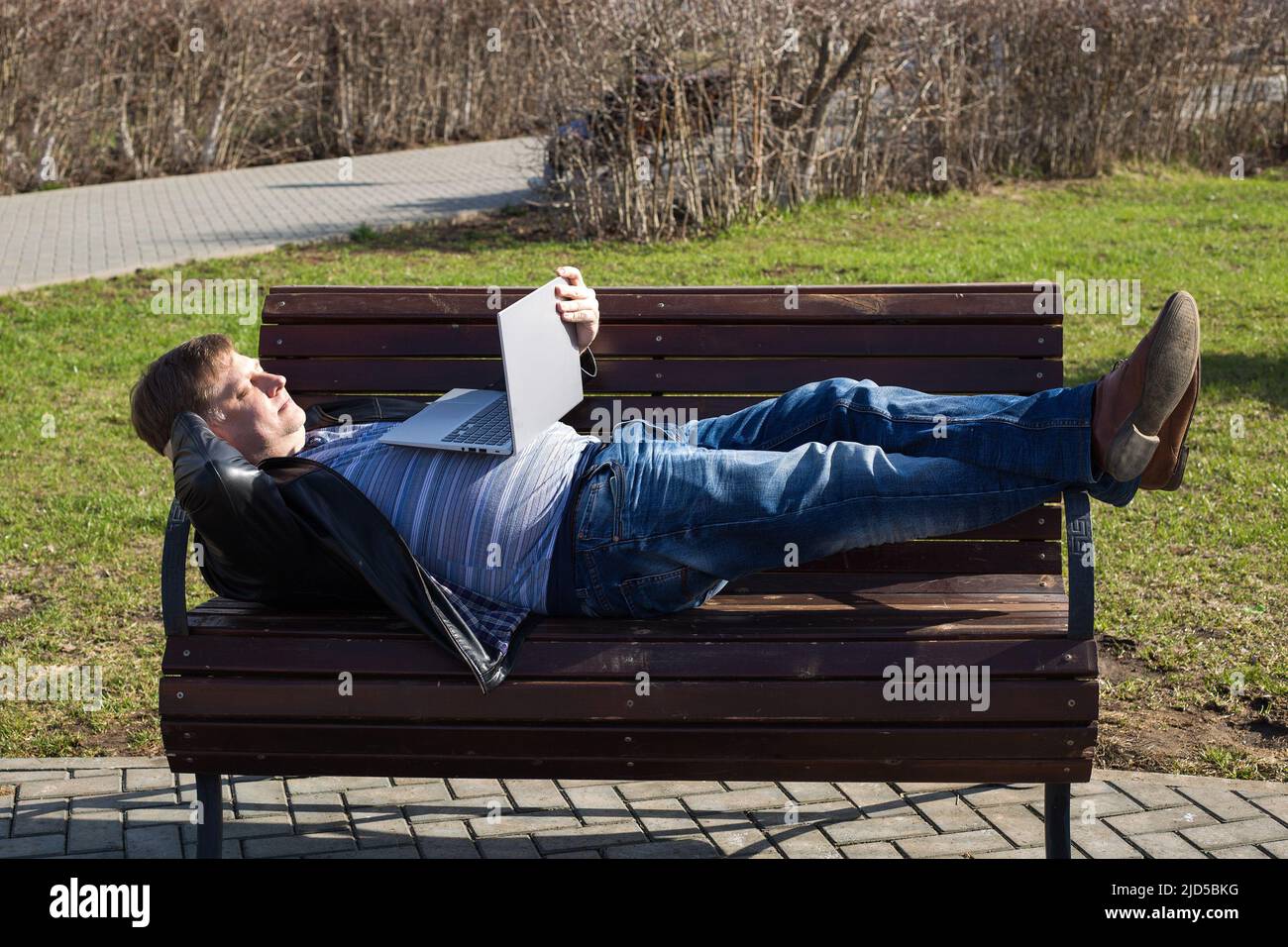 An adult man lay down on a bench in the park and watches a movie on a ...