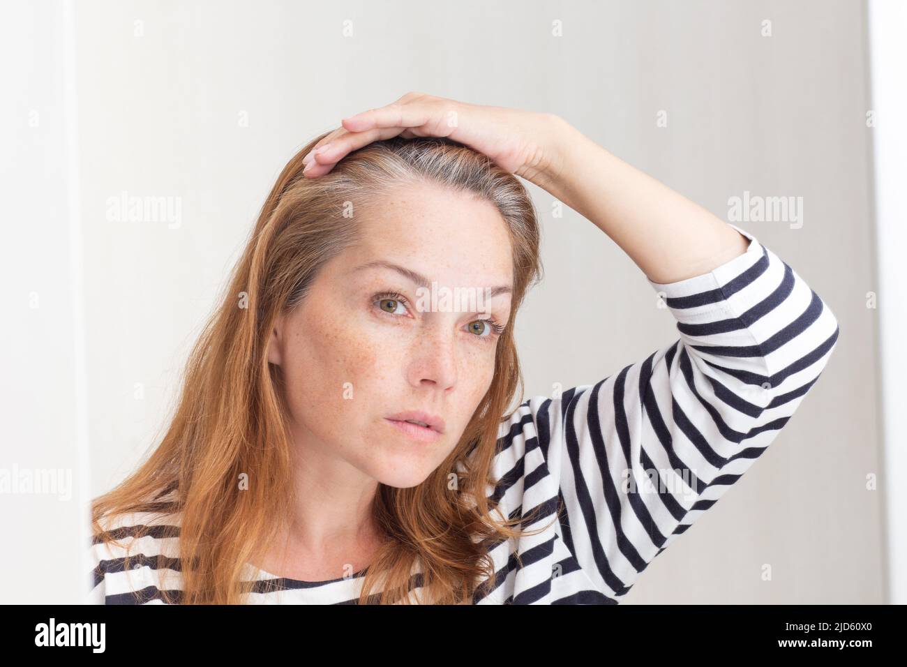 Serious 40s woman looking at gray hair in mirror reflection on growing ...