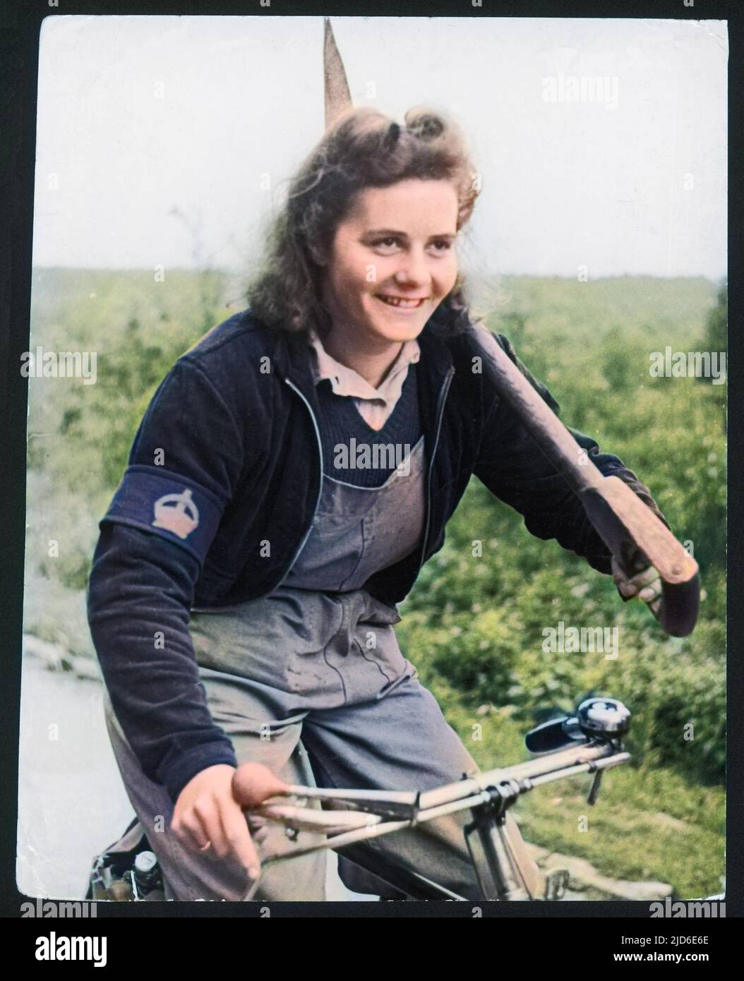 A cheerful Land Girl cycles to work with her spade over one shoulder ...