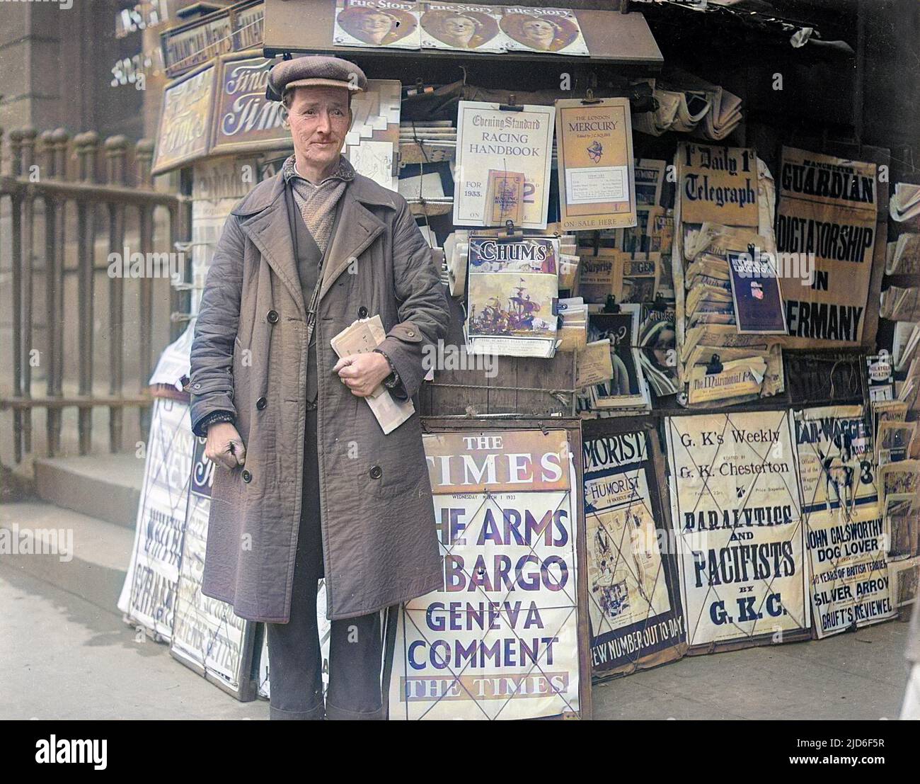 A newspaper seller in a flat cap and overcoat stands next to his stall ...