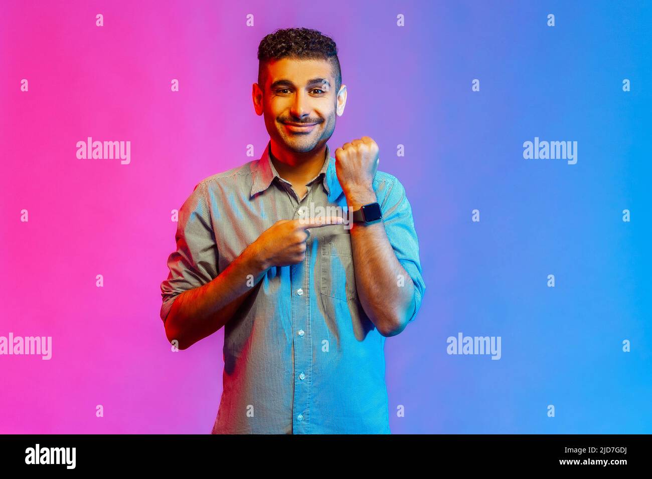 Portrait of cheerful punctual man in shirt pointing at wrist watch and ...