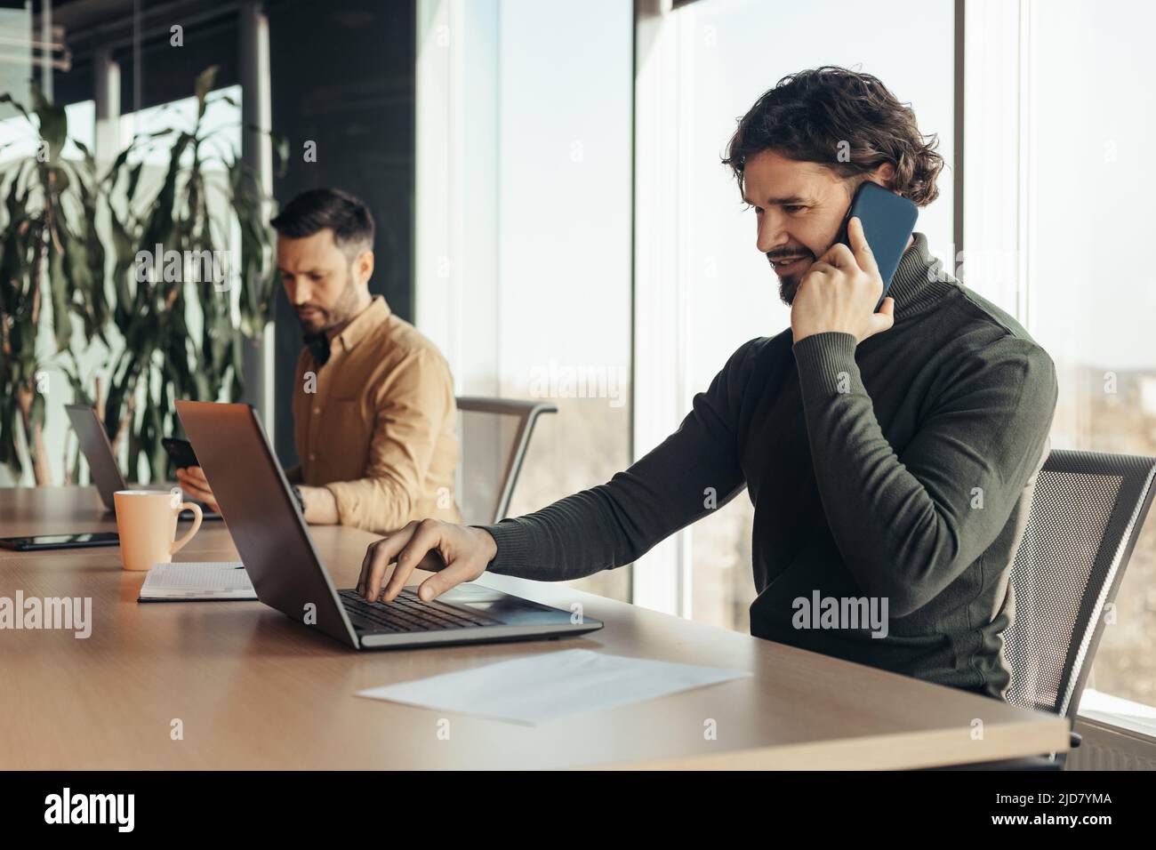 Joyful young businessman using laptop, talking on cellphone, working with male colleague at ...