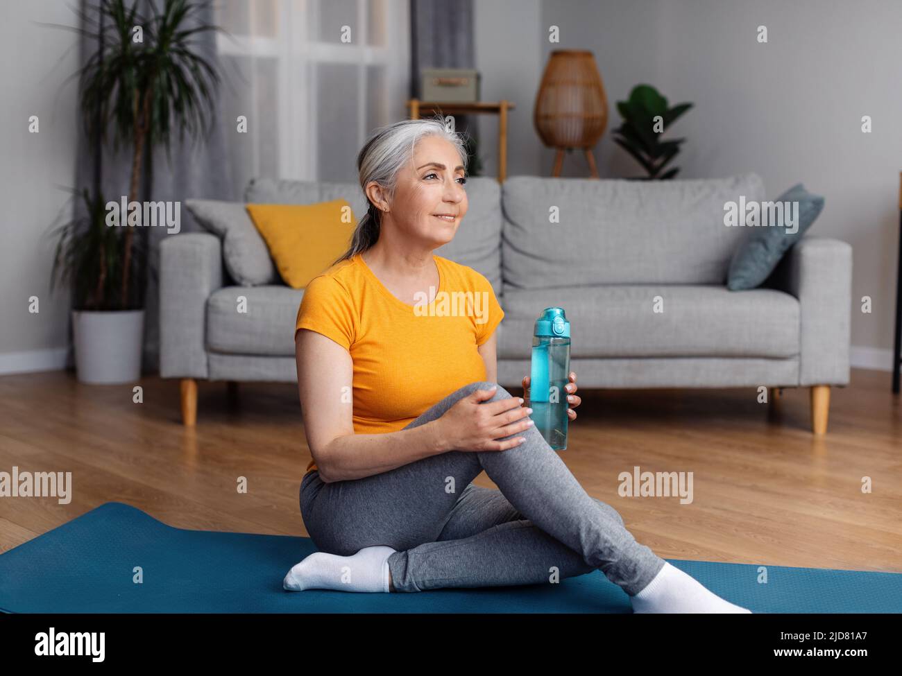 Positive sporty aged lady sitting on fitness mat with bottle of water ...