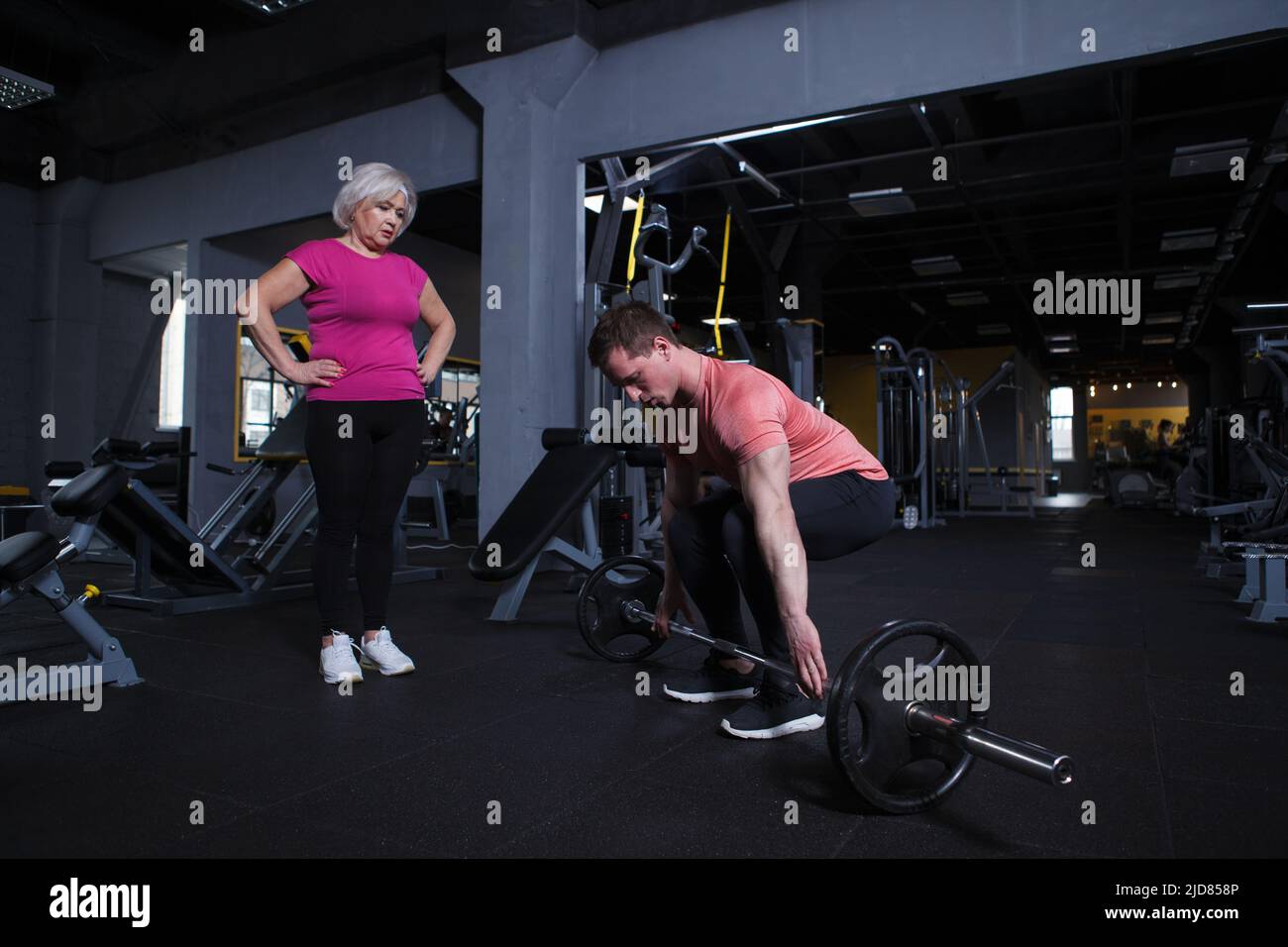 Athletic personal trainer showing his senior client how to lift barbell ...