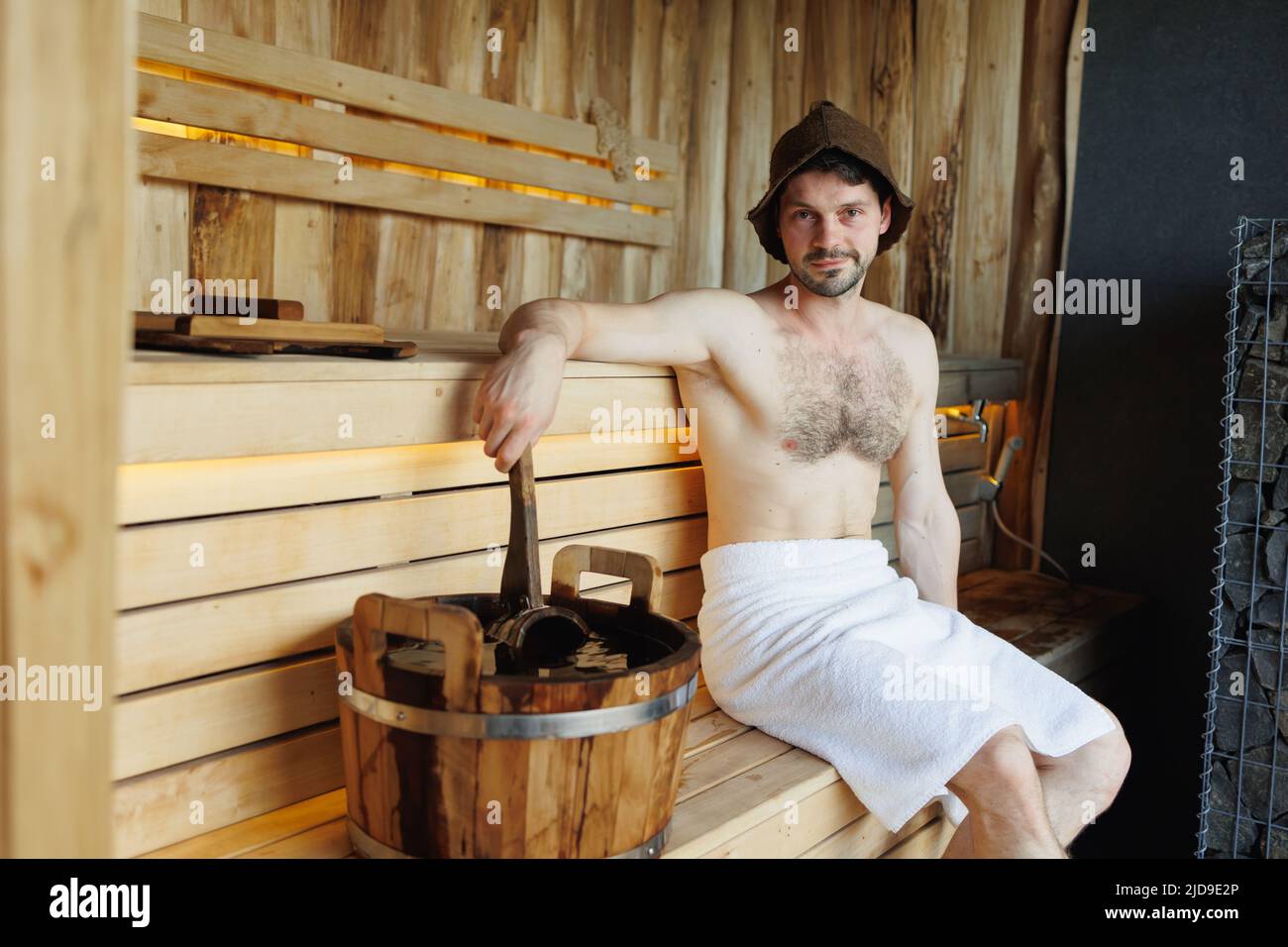 Handsome man sitting in the sauna bench takes water with a ladle from a ...