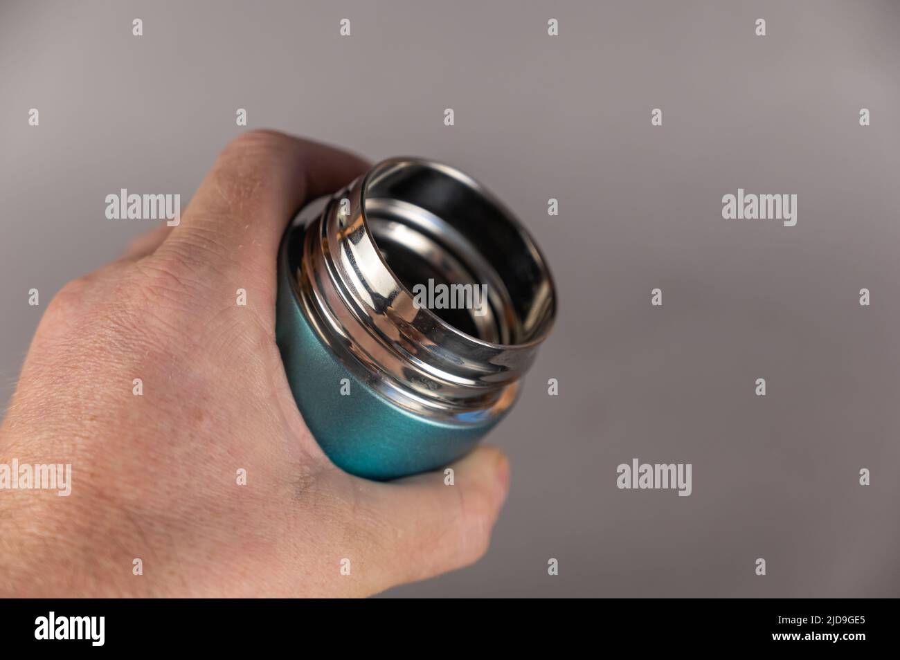 A man's hand holds an open metal thermos against a gray background. A ...