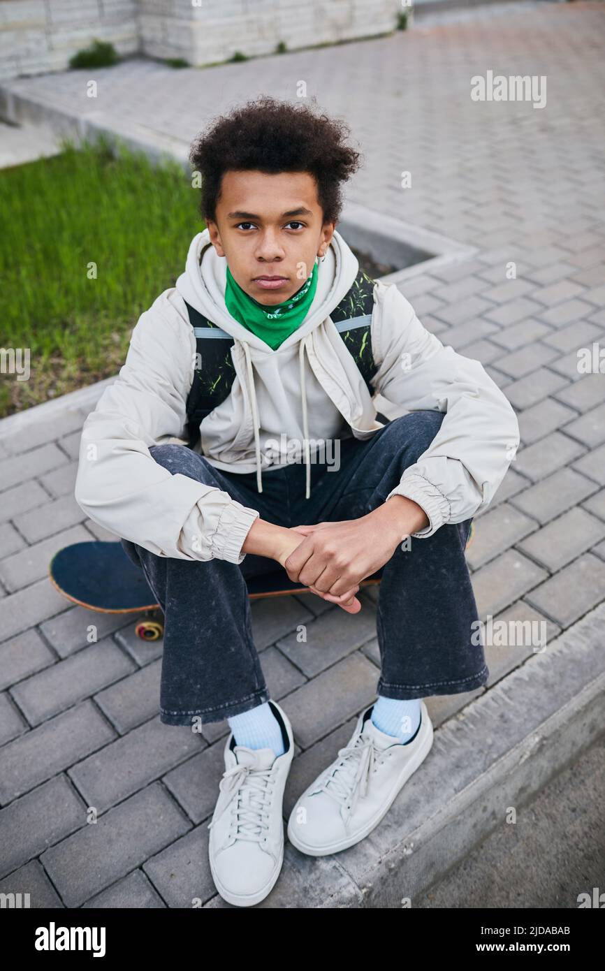 Portrait of African teenage boy sitting on his skateboard and looking at camera spending time ...