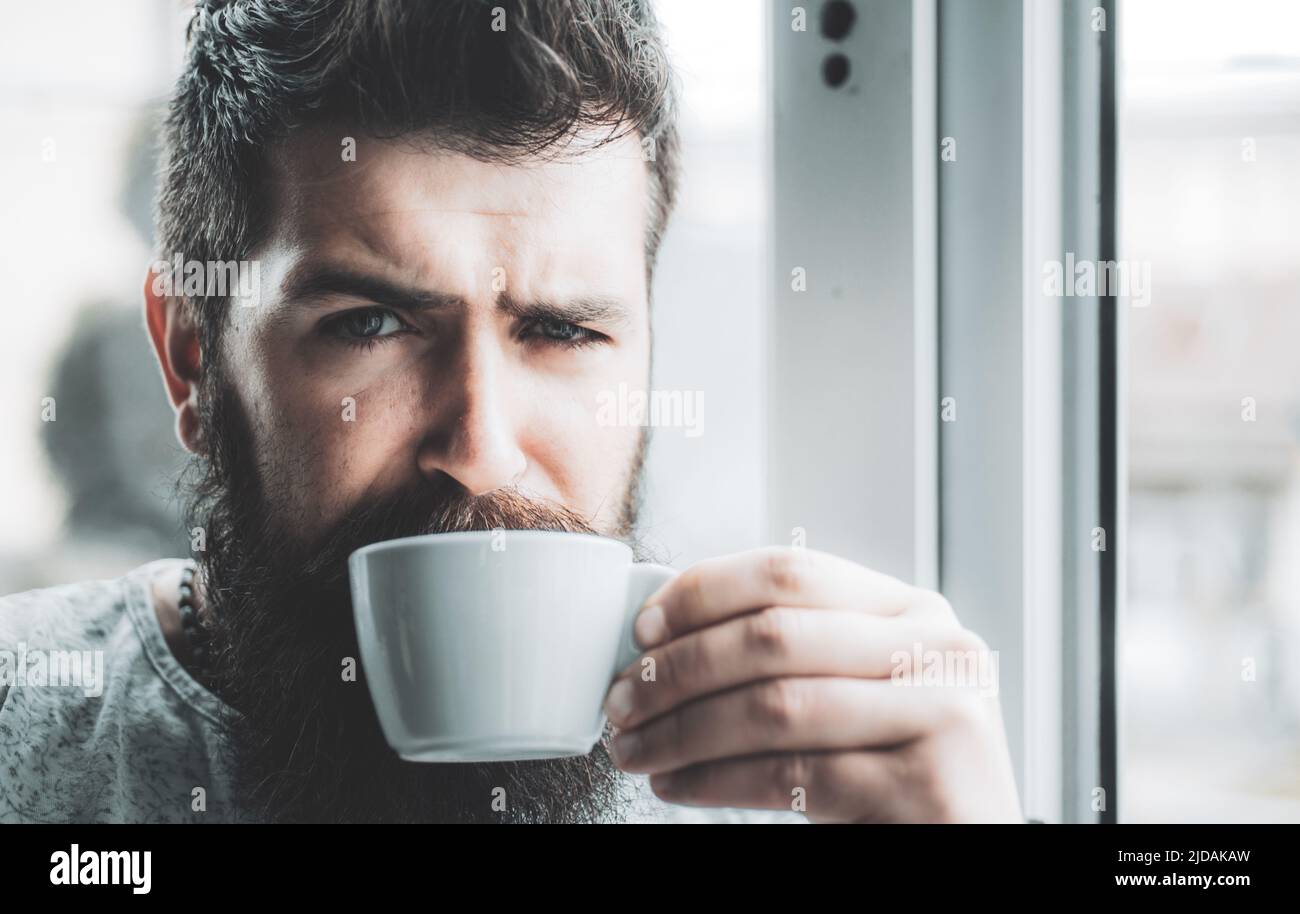 Grumpy looking bearded man drinking morning cup of coffee near window ...